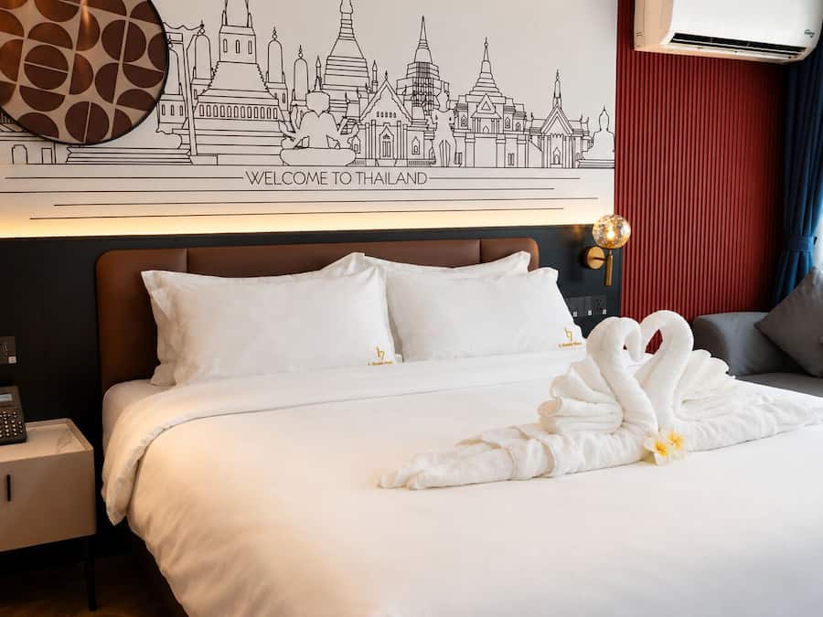 Quarto de hotel com cama, uma obra de arte na parede representando arquitetura tailandesa, arranjo de toalhas em forma de cisne e cortina vermelha.