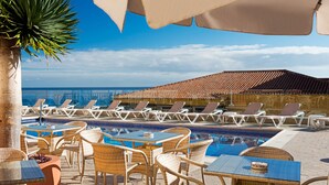 Uma área na piscina com espreguiçadeiras e mesas, vista desobstruída do oceano e uma cobertura com telhas.