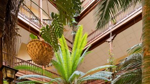 Um átrio espaçoso com vários níveis, destacando uma escadaria central, plantas tropicais exuberantes, móveis de vime e piso ladrilhado.