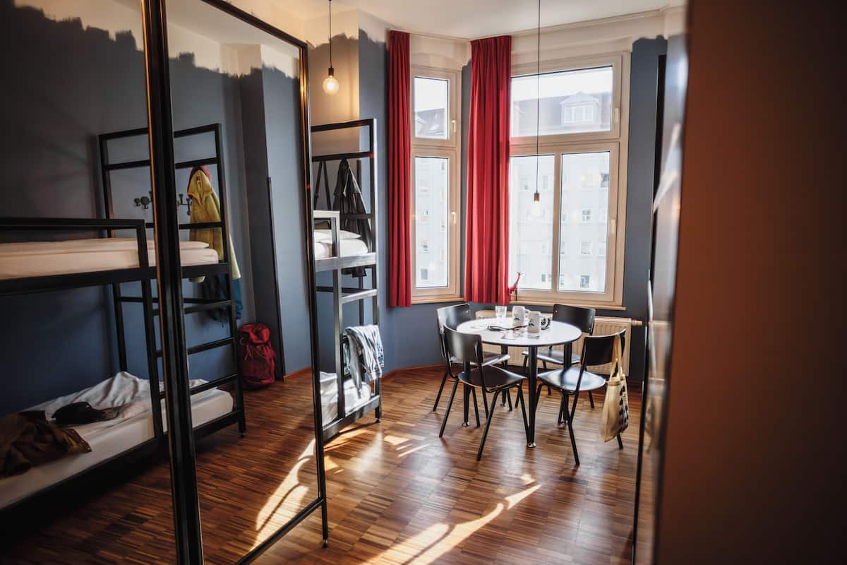 Quarto com beliches, mesa de jantar com cadeiras, janela com cortinas e radiador.