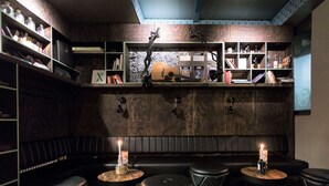 Uma área de bar com pouca luz, assentos de couro, um violão e prateleiras com livros e objetos decorativos.