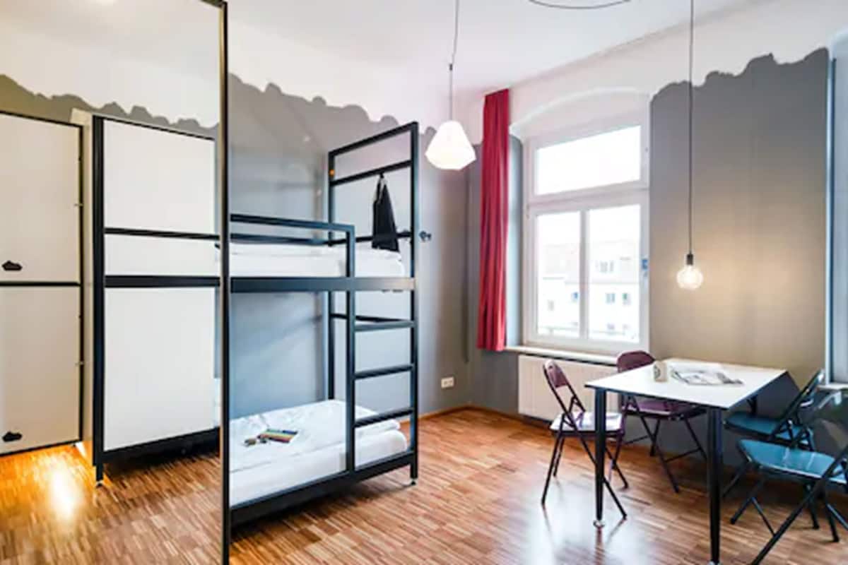 Um quarto com beliche, uma mesa de jantar com cadeiras e uma janela com cortinas vermelhas.