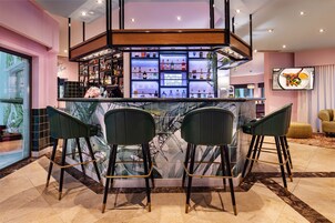 Uma área de bar com balcão de mármore, banquetas verdes e prateleiras com garrafas de bebidas.