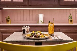 Uma mesa com um prato de comida, um copo alto de bebida e talheres. Há duas plantas em vasos na parede atrás da mesa.