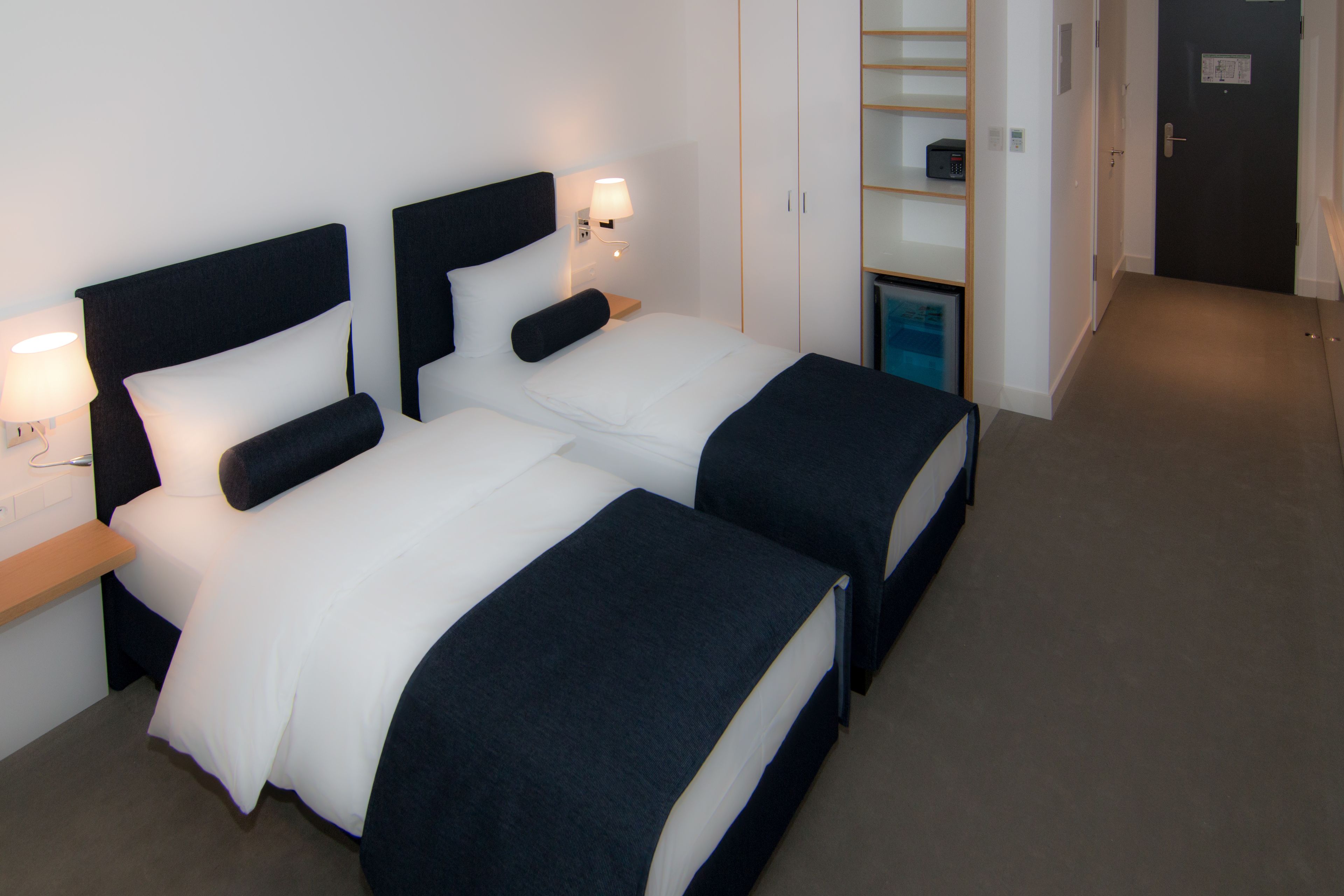 Quarto de hotel com duas camas, uma mesa de cabeceira com abajur, um guarda-roupa e uma pequena escrivaninha com computador.