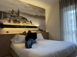 Quarto de hotel com cama, mesa de cabeceira, abajur e uma grande pintura na parede retratando uma cidade.