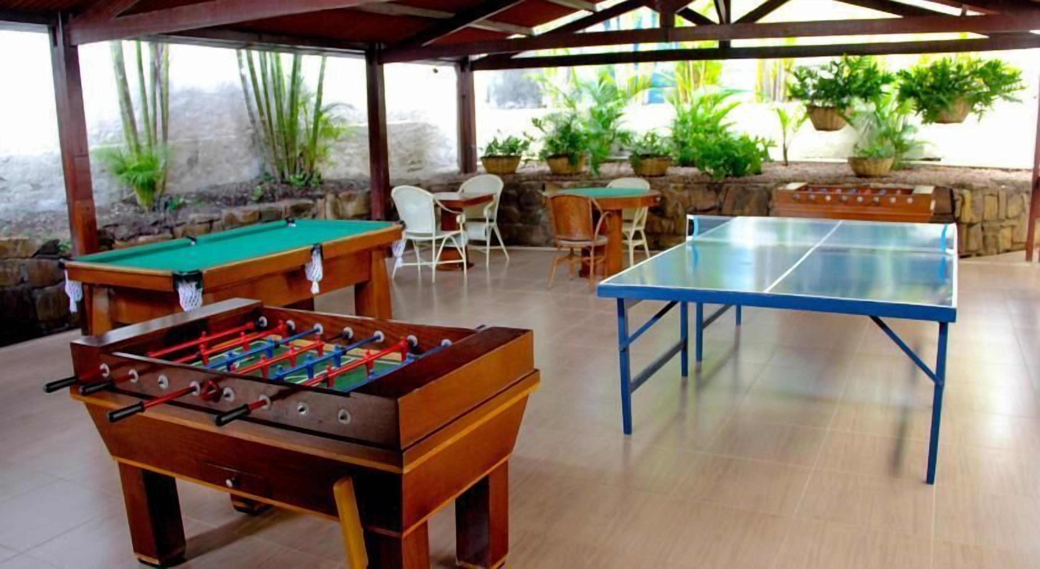 Mesa de bilhar, mesa de futebol de mesa e mesa de tênis em área externa coberta, com cadeiras e plantas em vasos.