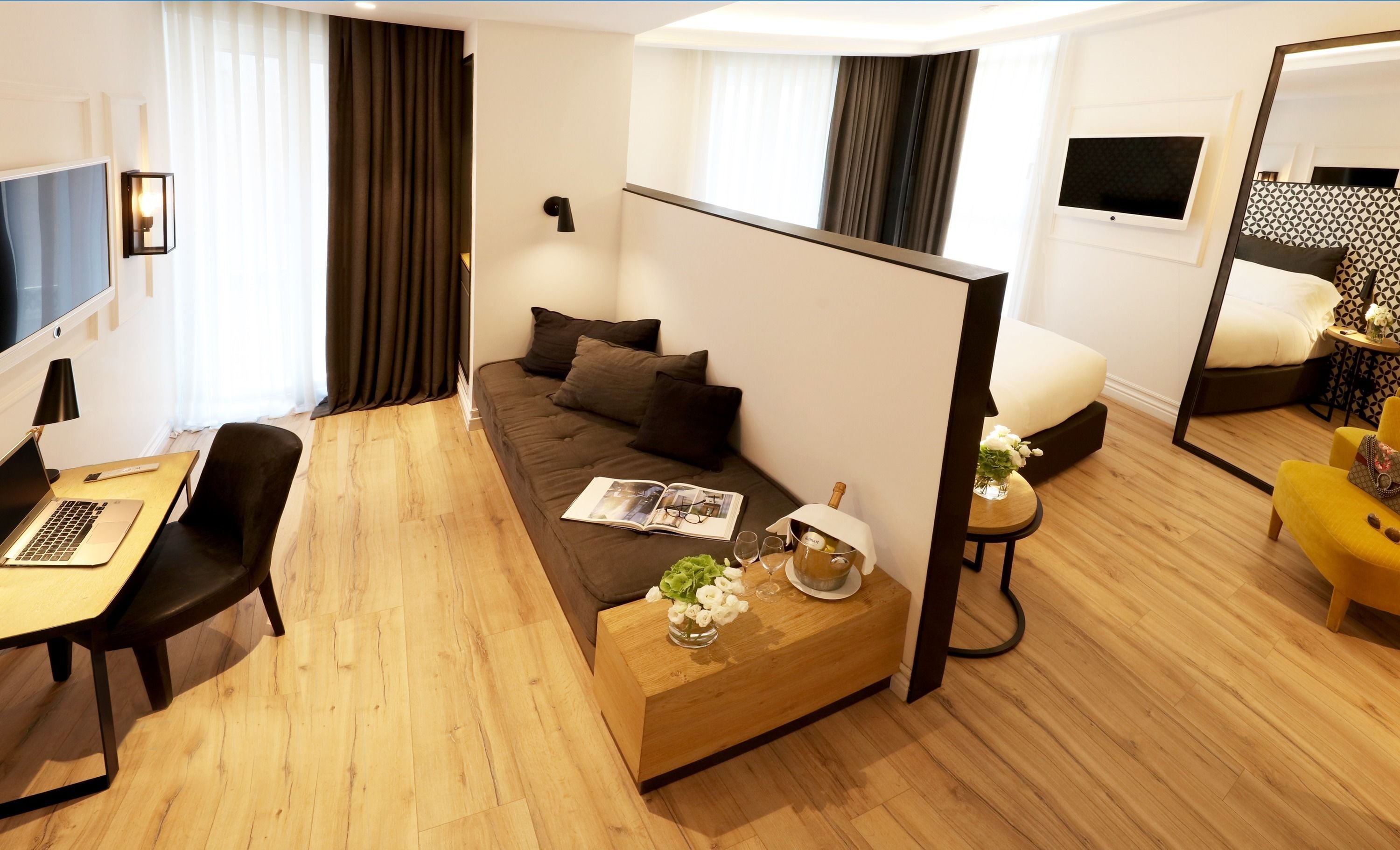 Quarto de hotel moderno com piso de madeira, um sofá, uma escrivaninha com um laptop, uma mesinha com uma garrafa e copos, e uma TV na parede.