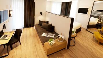 Quarto de hotel moderno com piso de madeira, um sofá, uma escrivaninha com um laptop, uma mesinha com uma garrafa e copos, e uma TV na parede.
