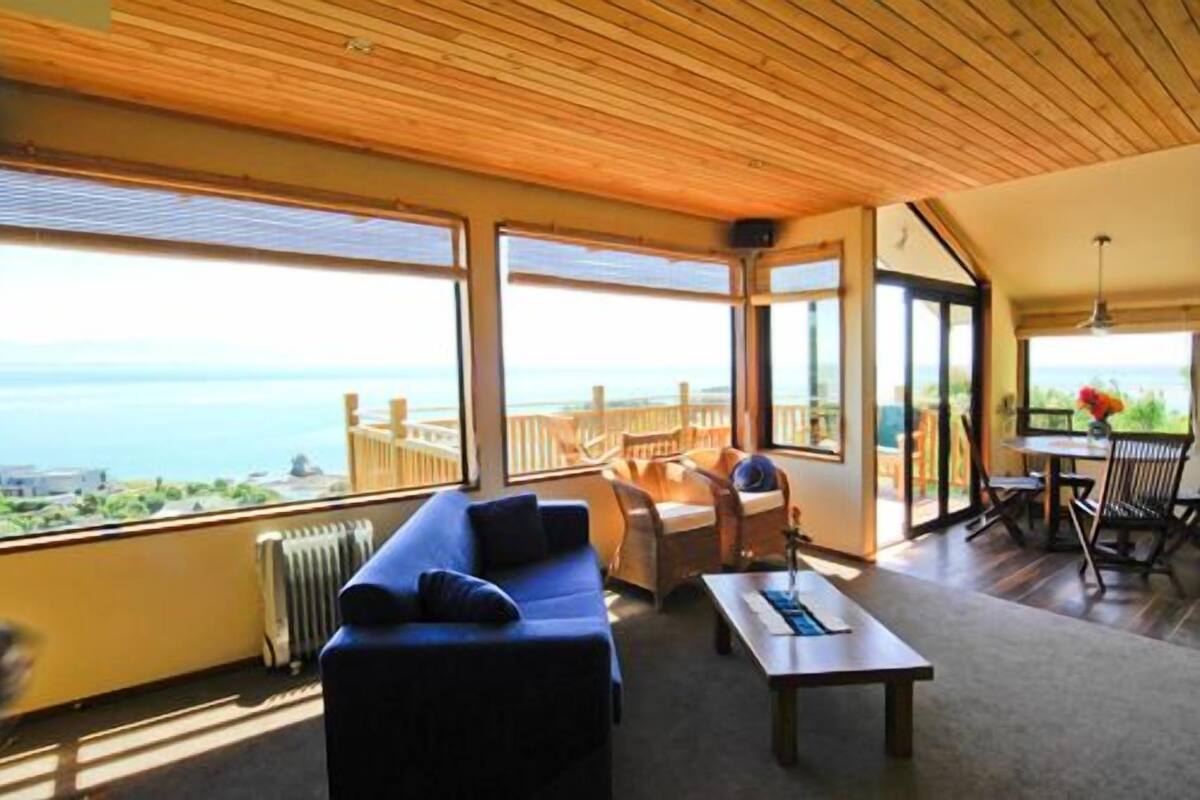 Quarto espaçoso com grande janela com vista para o mar, teto de madeira e área de estar acolhedora com sofá e mesa.
