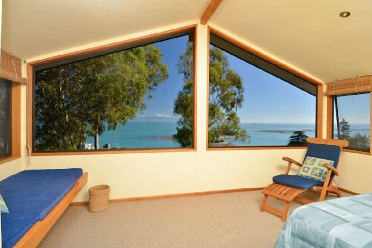 Quarto com uma grande janela que oferece vista para o mar e árvores. Há uma cama, uma cadeira e um banquinho de madeira.
