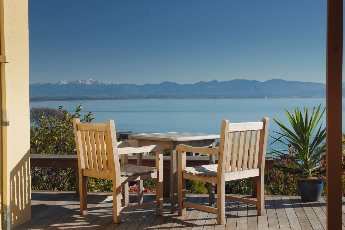 Um deck de madeira com mesa e cadeiras, oferecendo vista para um lago calmo e montanhas ao longe.