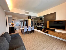 Uma sala de estar moderna com um sofá cinzento, um espaço de refeições com mesa e cadeiras, uma cozinha ao fundo e uma televisão de tela plana grande fixada na parede.