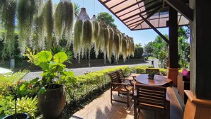 Uma área externa com mesas e cadeiras de madeira, rodeada por plantas em vasos e decorações com musgo pendurado.