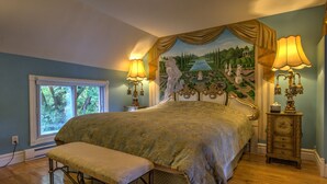 Quarto com uma cama grande, duas luminárias de cabeceira, um banco e uma janela com vista para árvores.
