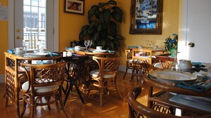 Uma sala de jantar com mesas e cadeiras de madeira, uma planta grande em vaso e um quadro na parede.