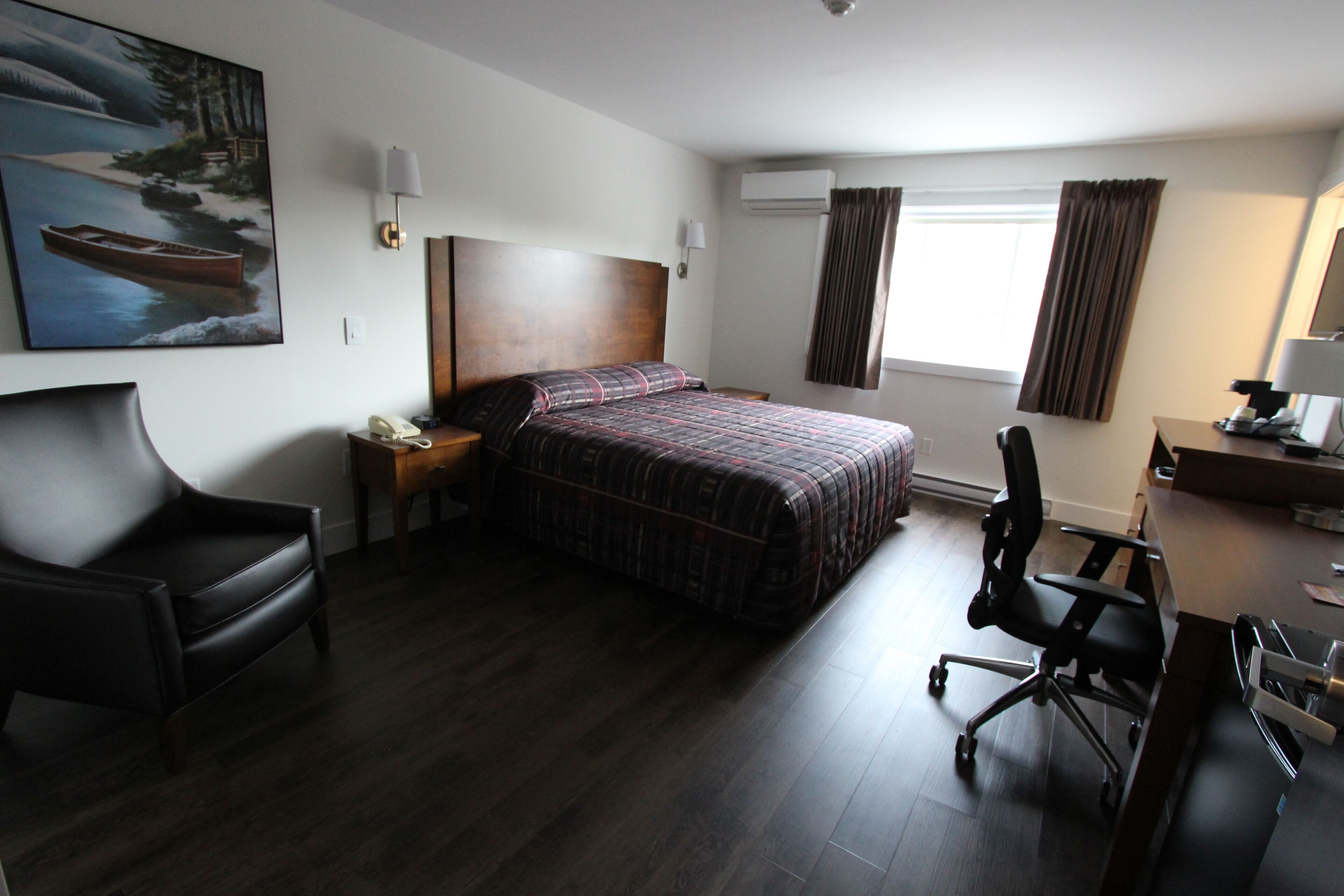 Quarto de hotel com cama, escrivaninha, cadeira, mesa de cabeceira, abajur e um quadro na parede.