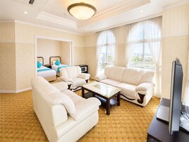 Quarto de hotel com sofá, poltronas, mesa de centro, televisão e cama com almofadas azuis.