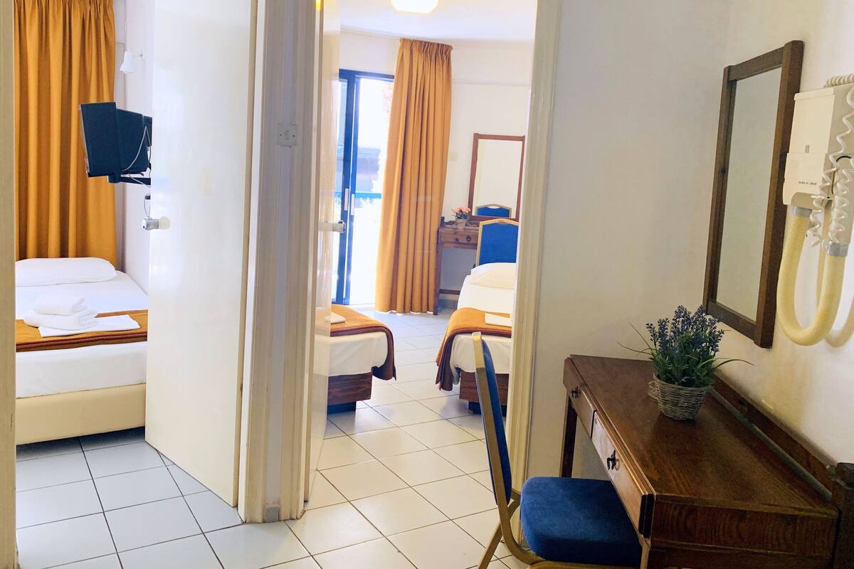 Quarto de hotel com duas camas, uma escrivaninha com cadeira, um espelho e uma planta.