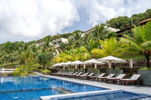Uma área de piscina com espreguiçadeiras e guarda-sóis, cercada por vegetação exuberante e uma encosta.