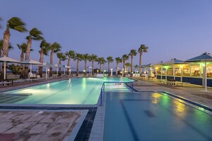 Piscina com placa de 'Não Mergulhar', rodeada por palmeiras e espreguiçadeiras, com vista para o oceano ao entardecer.