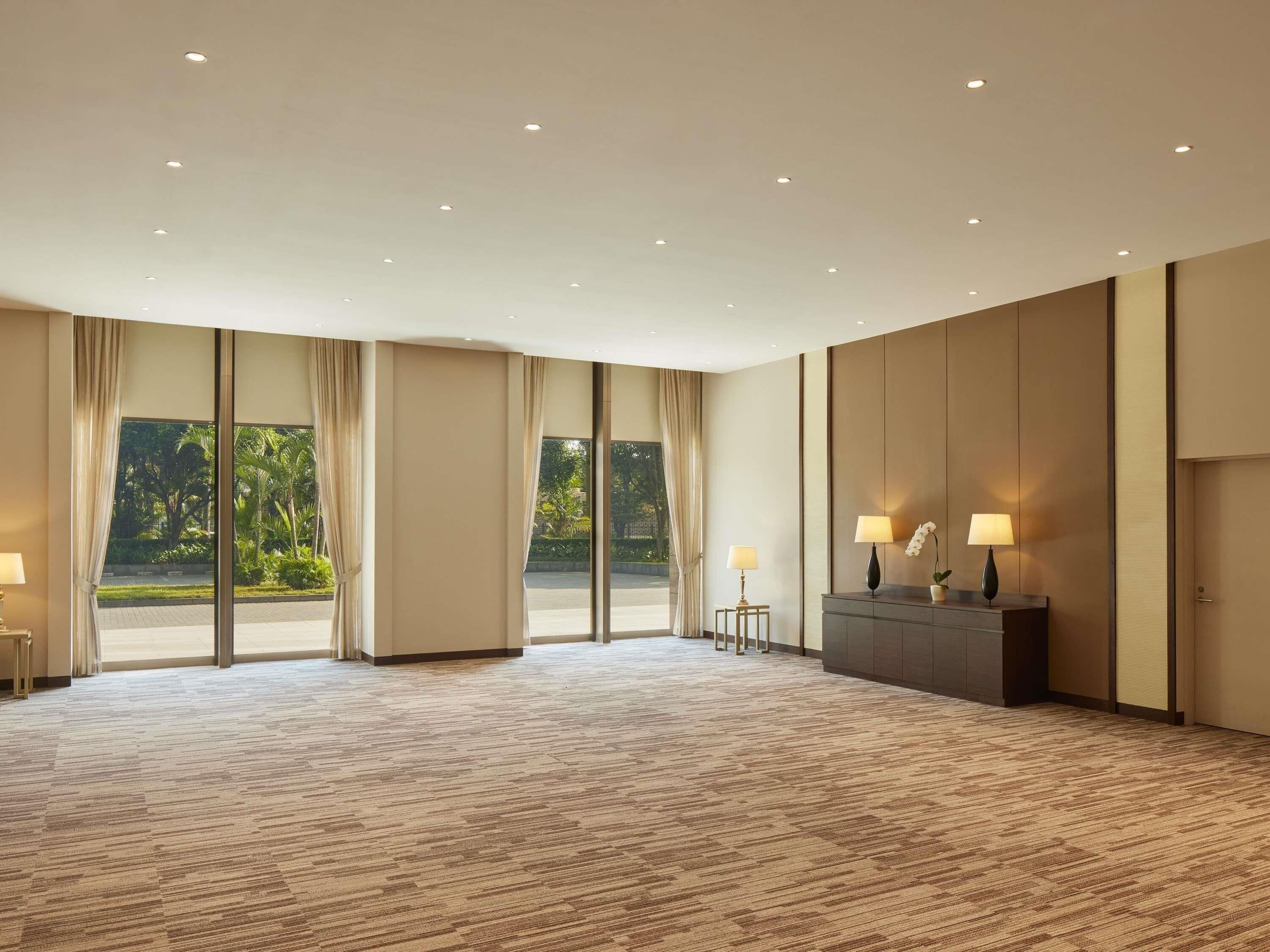 Quarto de hotel espaçoso com janelas amplas, piso carpetado, armário de madeira com luminárias e vista para o verde lá fora.