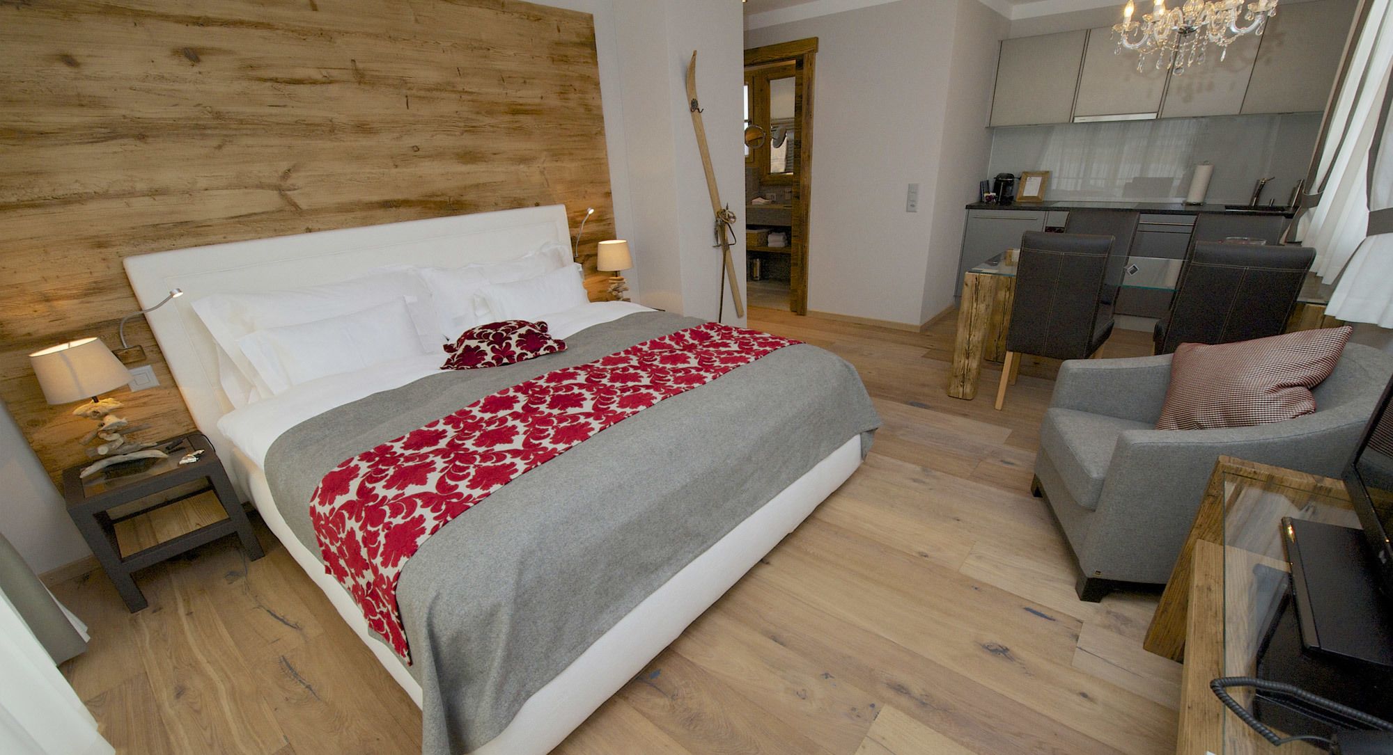 Quarto com cama grande, mesa de cabeceira com abajur, cabeceira de madeira e sofá cinza com almofada.