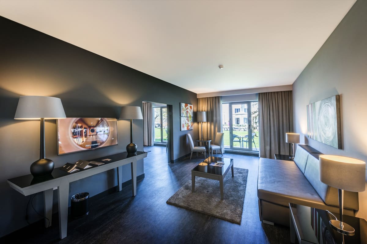 Quarto de hotel moderno com sofá escuro, mesa de centro, TV fixada na parede e uma grande janela com cortinas.