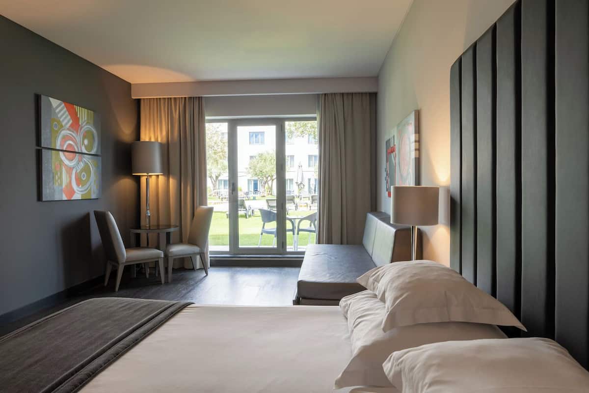 Quarto de hotel com cama, escrivaninha, cadeira, luminária e vista para o exterior.