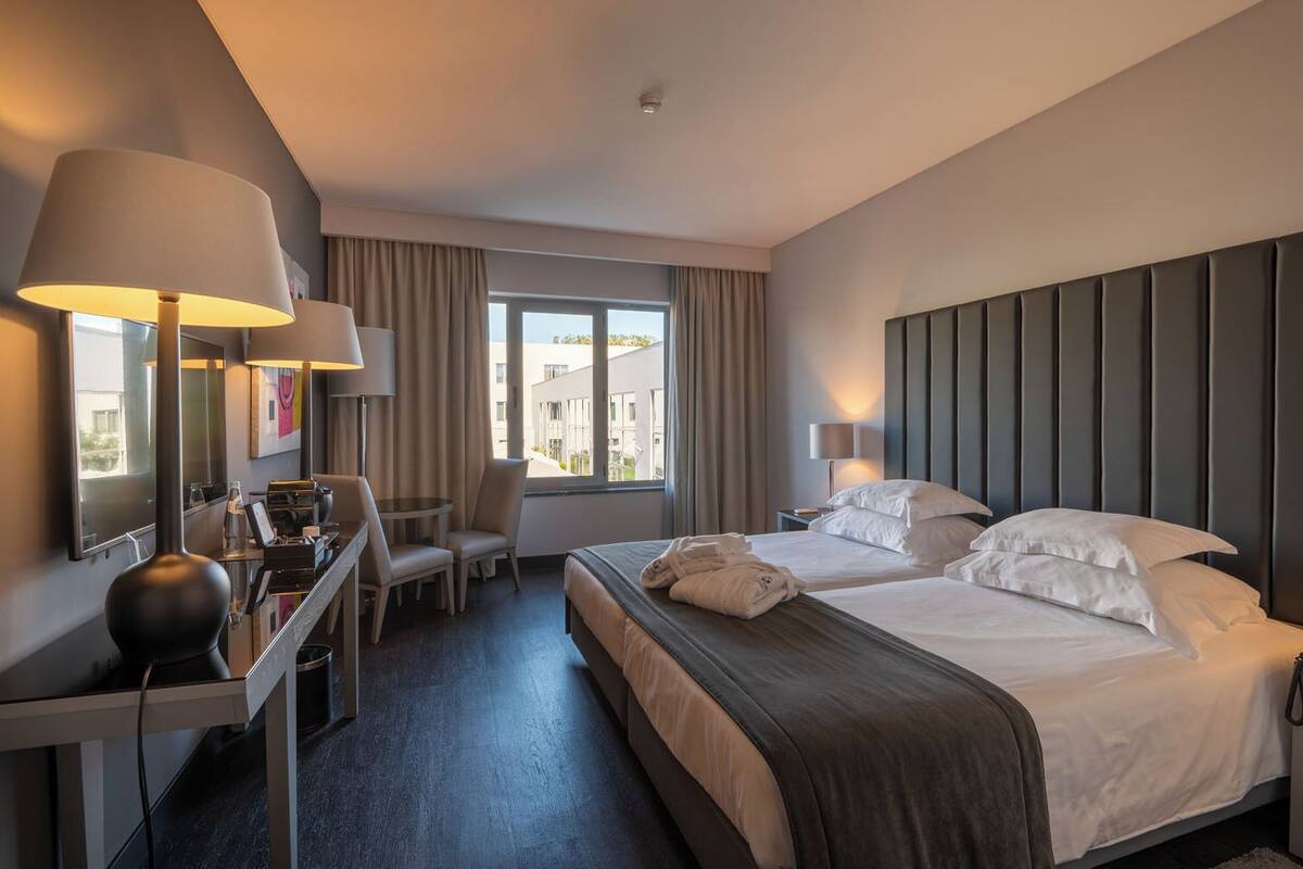 Quarto de hotel com cama, mesas de cabeceira, escrivaninha, cadeira e janela com cortinas.