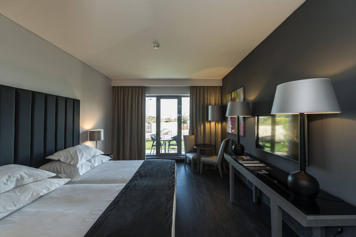 Quarto de hotel moderno com uma cama grande, uma escrivaninha com luminária, uma cadeira e vista para o exterior.