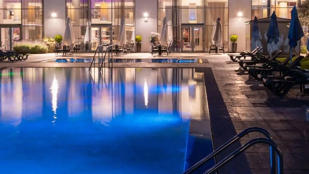 Piscina com espreguiçadeiras e guarda-sóis, cercada por um edifício com grandes janelas e interior bem iluminado.
