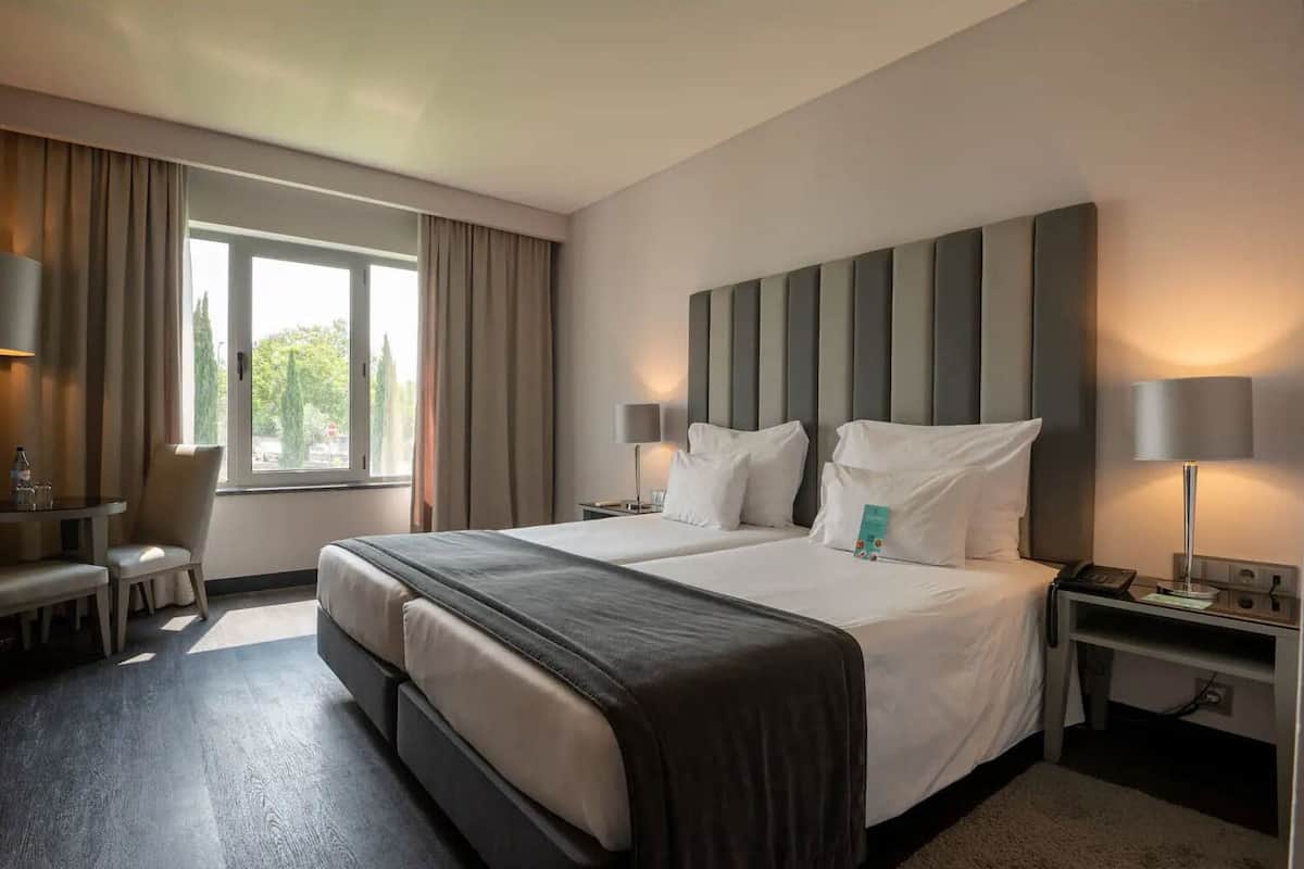 Quarto de hotel com uma cama grande, mesas de cabeceira, uma escrivaninha com cadeira e uma janela com cortinas.
