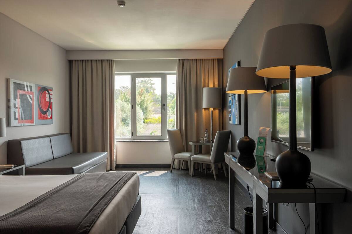 Quarto de hotel com cama, escrivaninha com luminária, cadeira, sofá, janela com cortinas e obras de arte na parede.