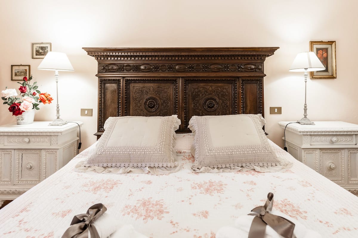 Cama bem arrumada com colcha florida, dois travesseiros e um painel de cabeceira decorativo. Duas mesas de cabeceira, com luminárias e um vaso de flores sobre uma delas.