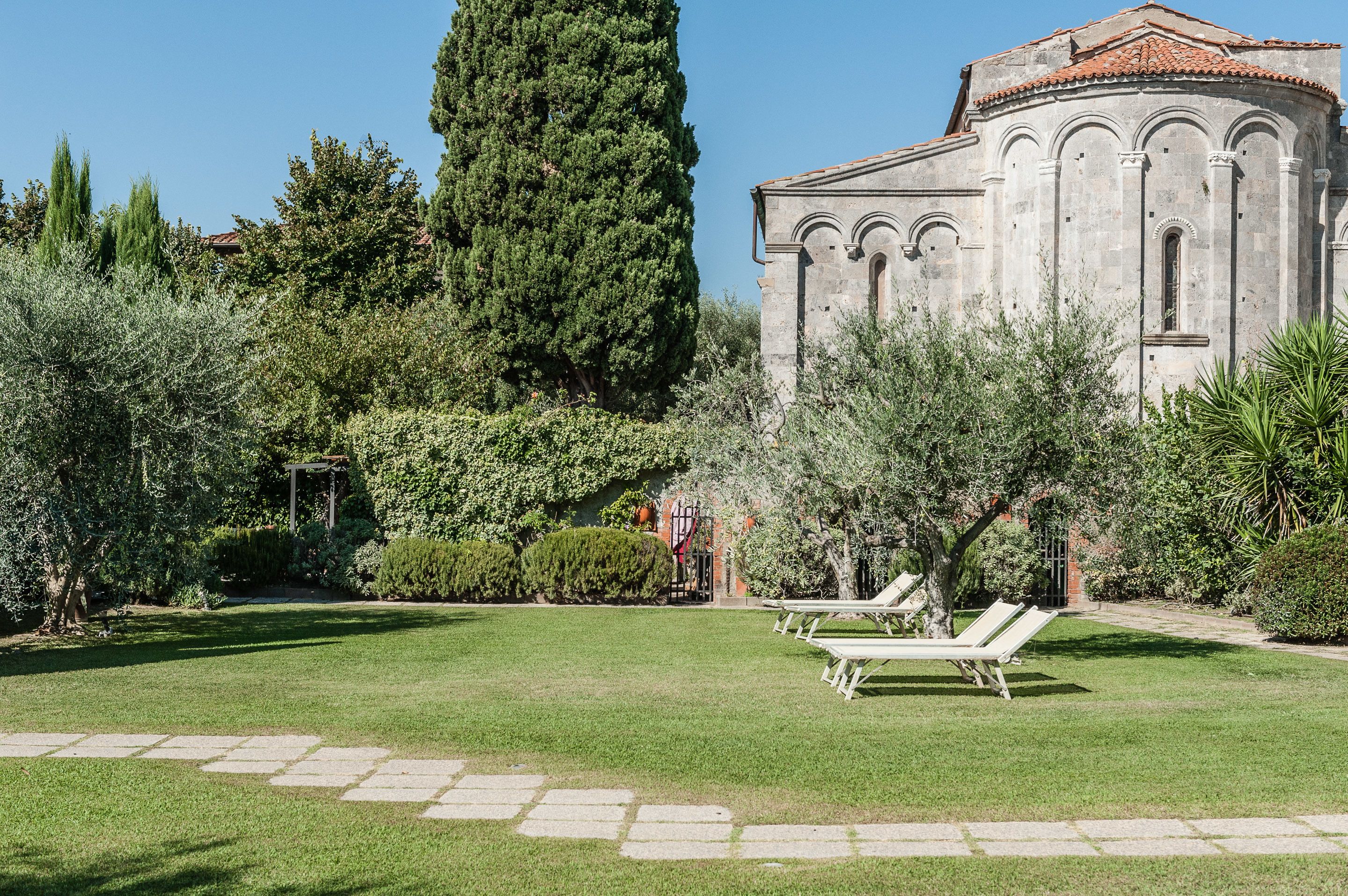 Um pátio com um edifício com janelas em arco e telhado de azulejos, cercado por vegetação exuberante e um caminho de pedra.