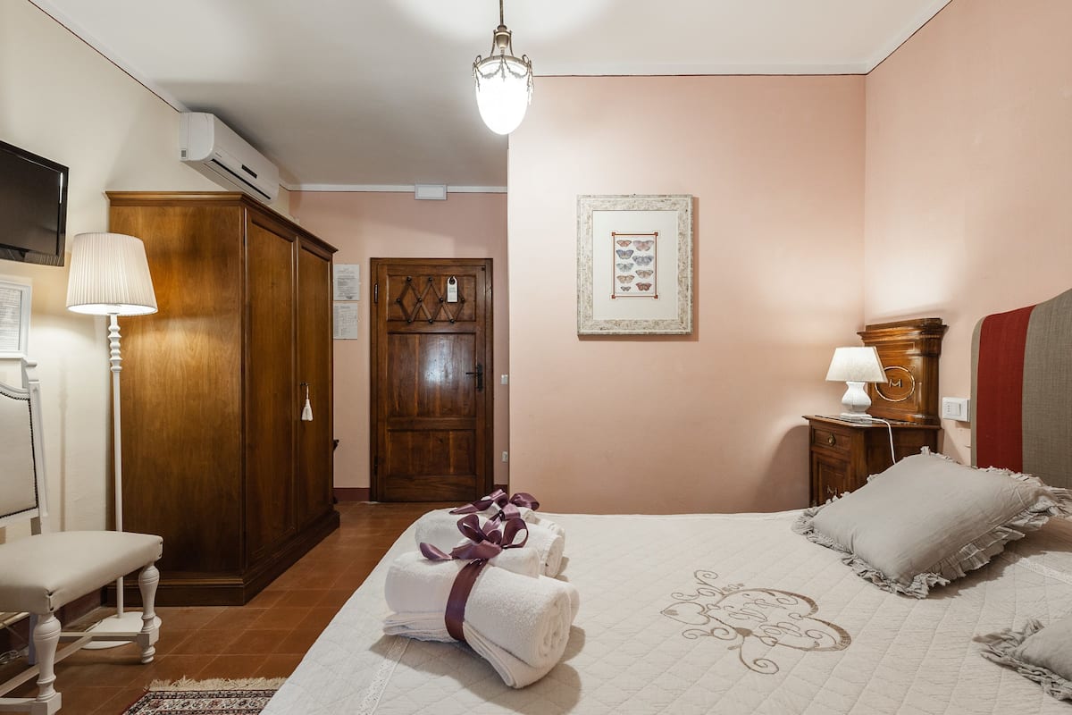Quarto de hotel com uma cama, um guarda-roupa de madeira, uma escrivaninha com abajur e um quadro na parede.