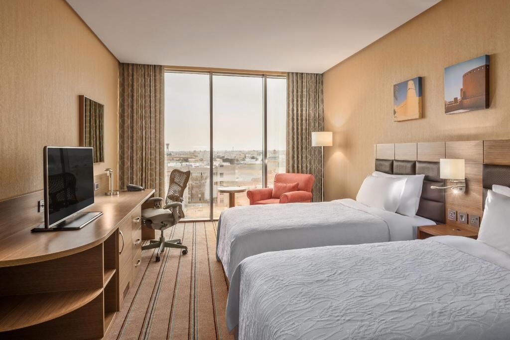 Quarto de hotel com duas camas, uma mesa de escritório com televisão, uma cadeira, uma mesinha e vista da cidade através de grandes janelas.