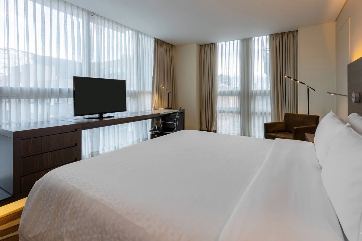 Quarto de hotel com uma cama grande, uma escrivaninha com televisão de tela plana, uma cadeira e uma luminária de mesa.