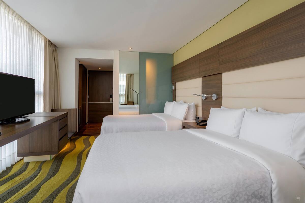 Quarto de hotel com duas camas, uma mesa de trabalho com televisão e vista para o corredor.