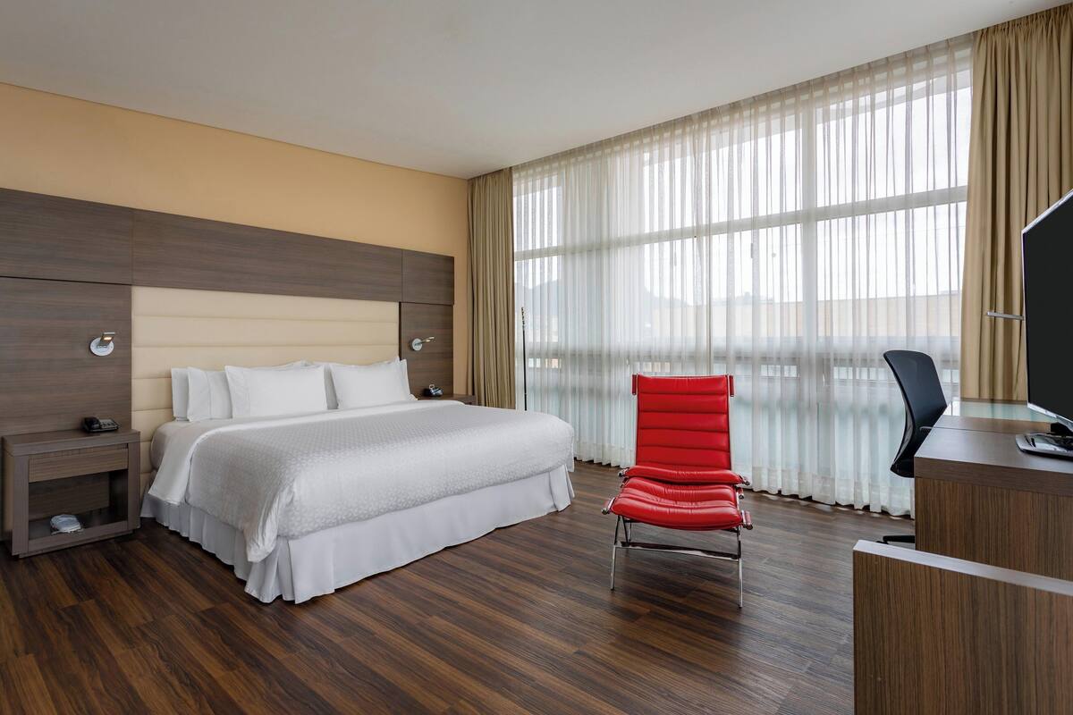 Quarto de hotel com uma cama grande, uma cadeira vermelha, uma mesa com computador e uma janela ampla com cortinas translúcidas.