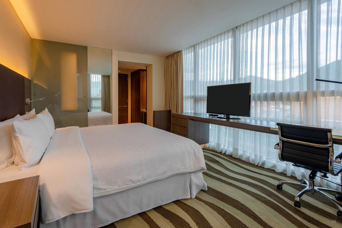 Quarto de hotel com uma cama grande, uma escrivaninha com televisão de tela plana, uma cadeira e vista para o exterior através de cortinas finas.