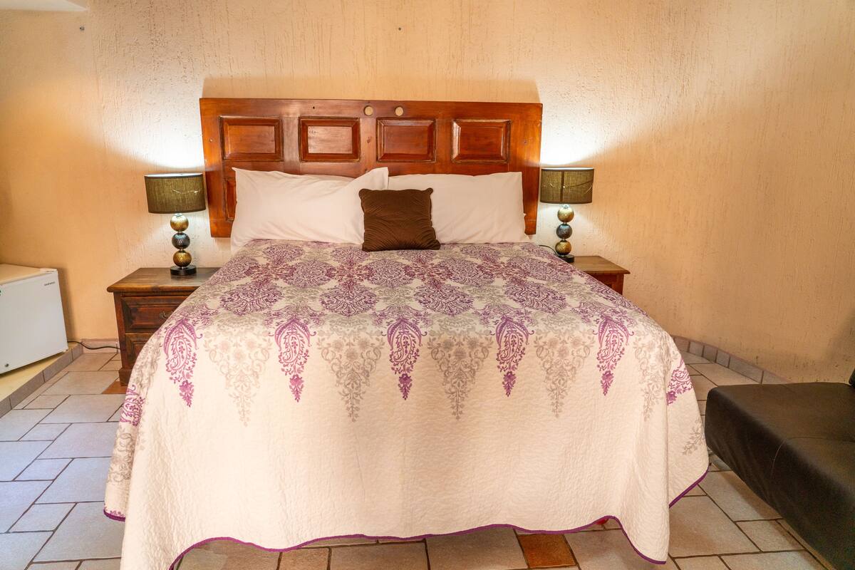 Cama bem arrumada com colcha estampada, cabeceira de madeira e duas luminárias laterais.