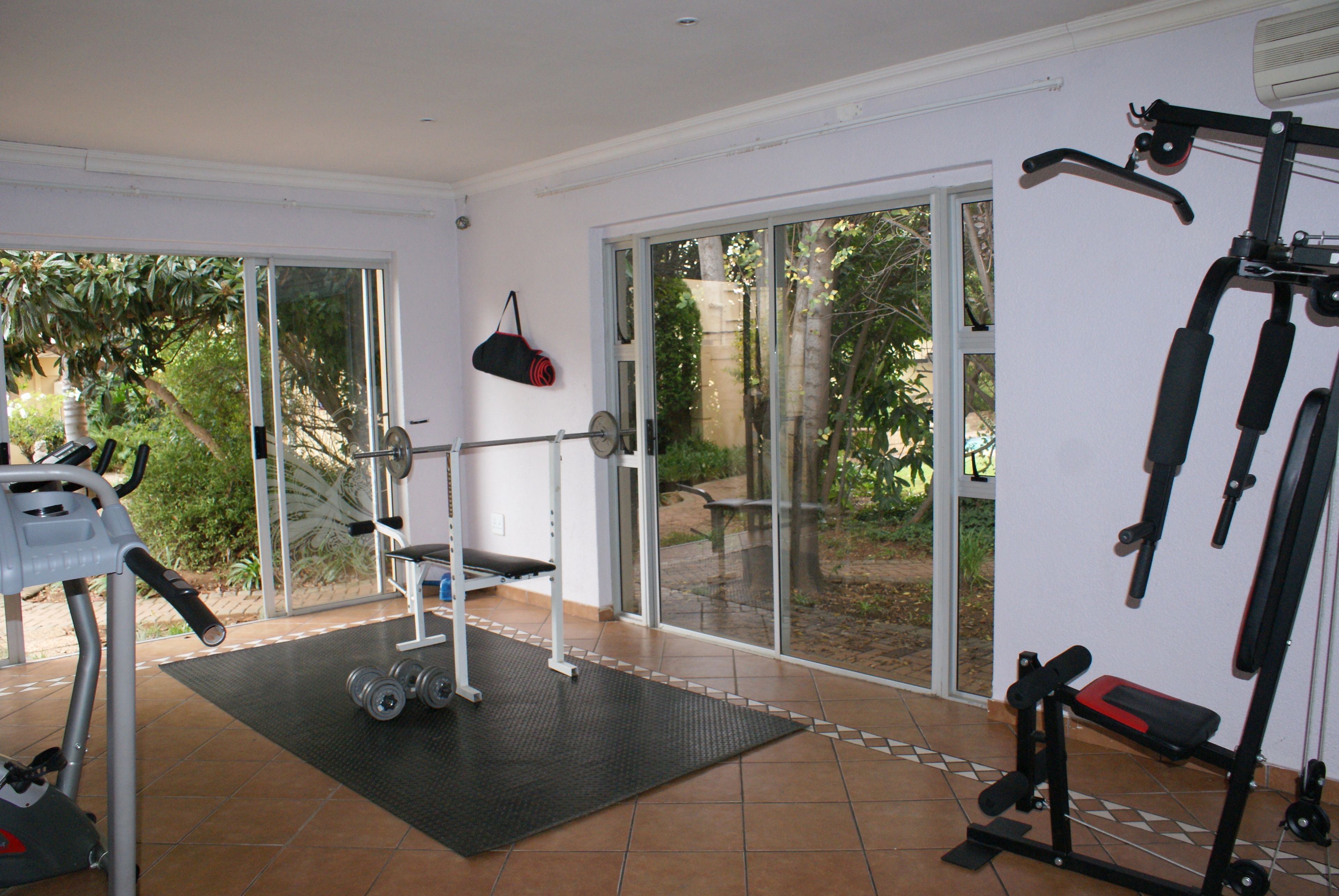 Uma academia em casa com equipamentos de exercício, uma porta de correr com vidro que dá para um jardim e um tapete no chão.