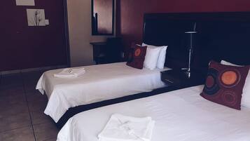 Quarto de hotel com duas camas, cada uma com lençóis brancos e almofadas decorativas. Há uma mesinha de cabeceira com uma luminária e um espelho na parede.