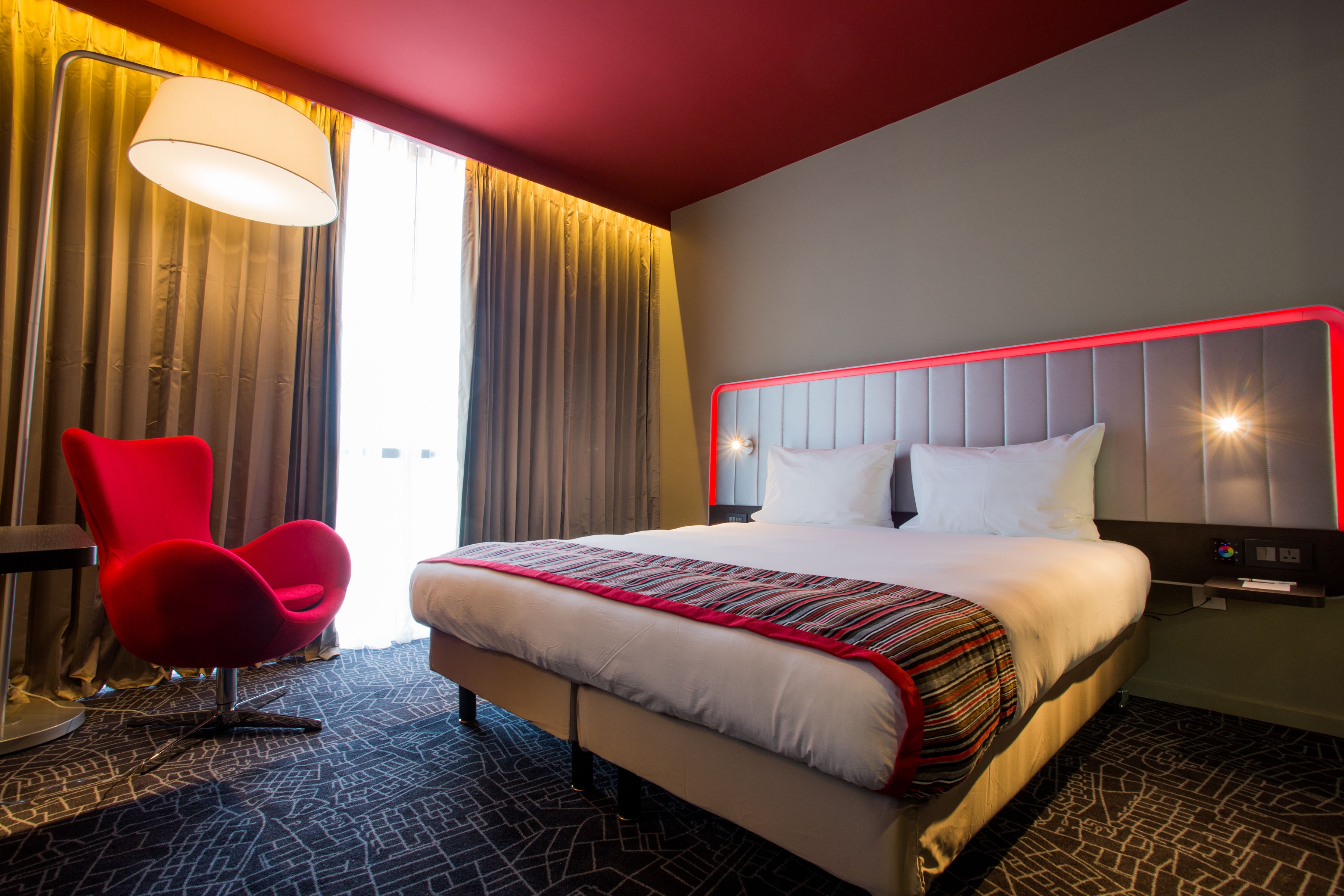 Quarto de hotel com uma cama grande, uma cadeira vermelha, uma escrivaninha e uma luminária.