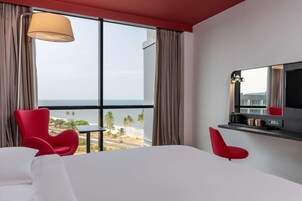 Quarto de hotel com uma cama grande, duas cadeiras vermelhas, uma TV em uma estante e vista para o mar.