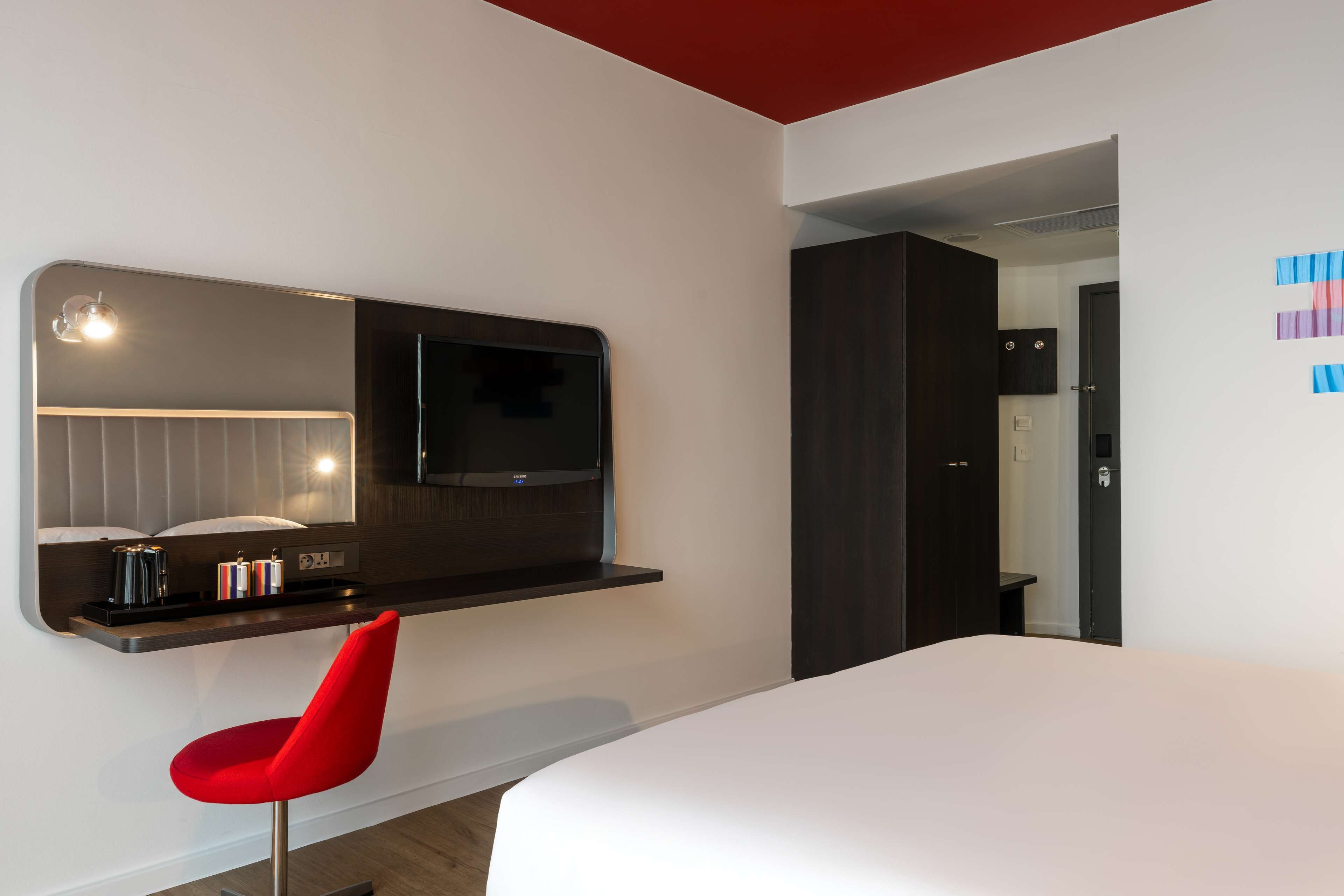 Quarto de hotel moderno com uma cama grande, uma cadeira vermelha, uma televisão de tela plana fixada em uma parede escura e uma pequena mesa com livros e uma luminária.