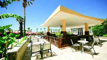 Uma área de pátio ao ar livre com mesas e cadeiras, cercada por palmeiras e plantas em vasos.
