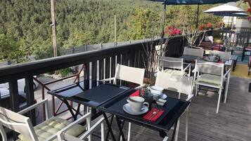 Um terraço ao ar livre com mesas, cadeiras, guarda-sóis e vista para uma área arborizada.